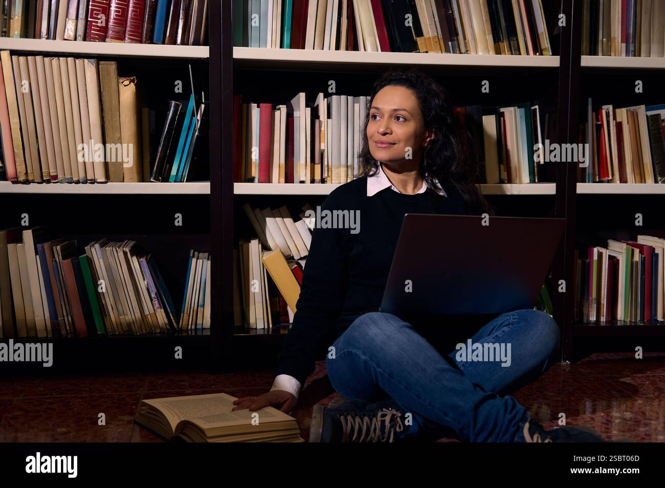 A woman sitting comfortably on the floor of a library, using a laptop ...