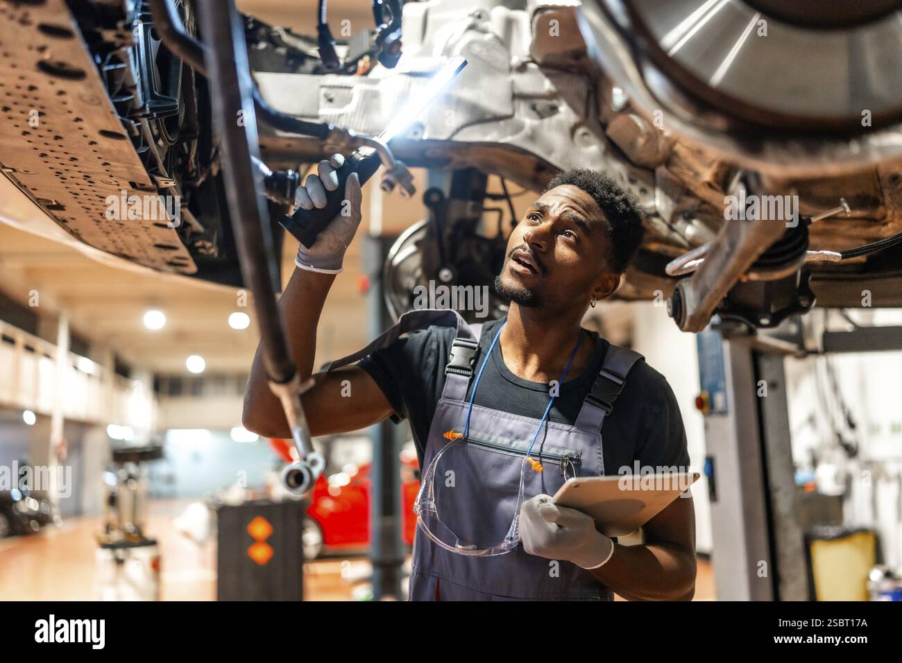 Young mechanic inspecting car chassis using flashlight and holding ...
