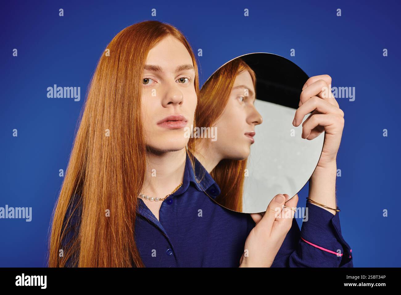 A young queer man with long red hair looks at himself in a circular ...