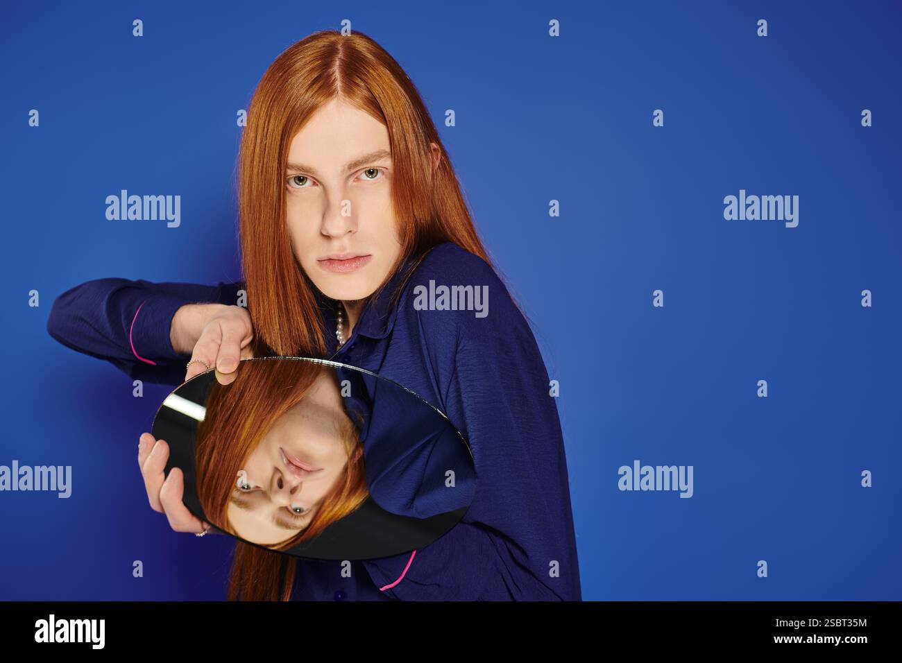 A young queer man with long red hair strikes a bold pose with a mirror ...