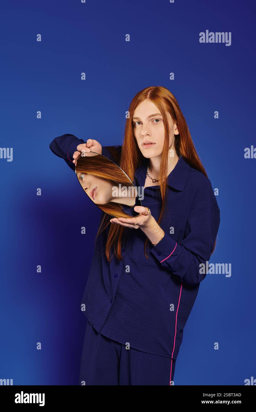 A young queer man with long red hair holds a mirror, his face ...