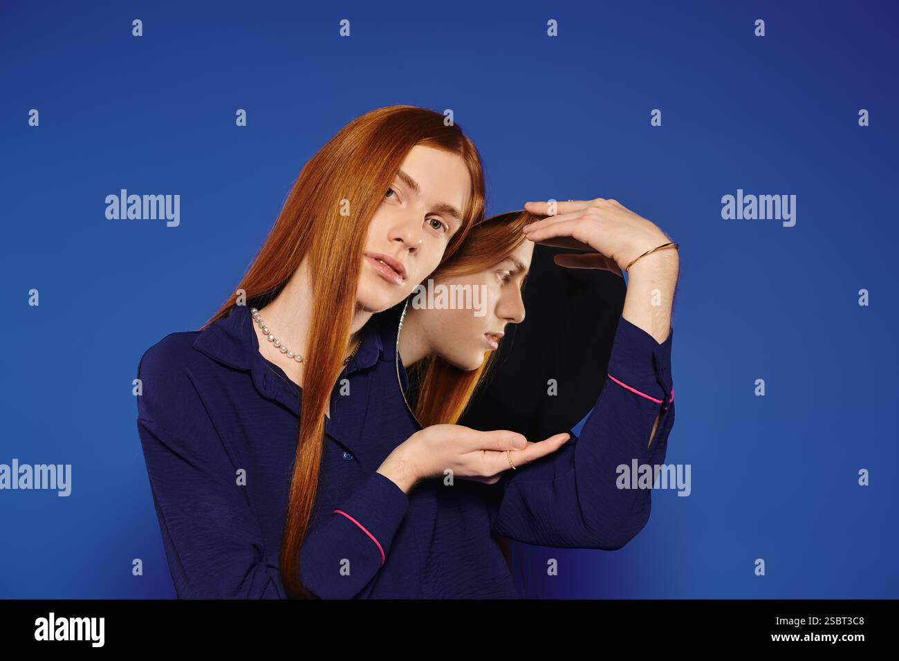 A young queer man with long red hair gazes thoughtfully into a mirror ...