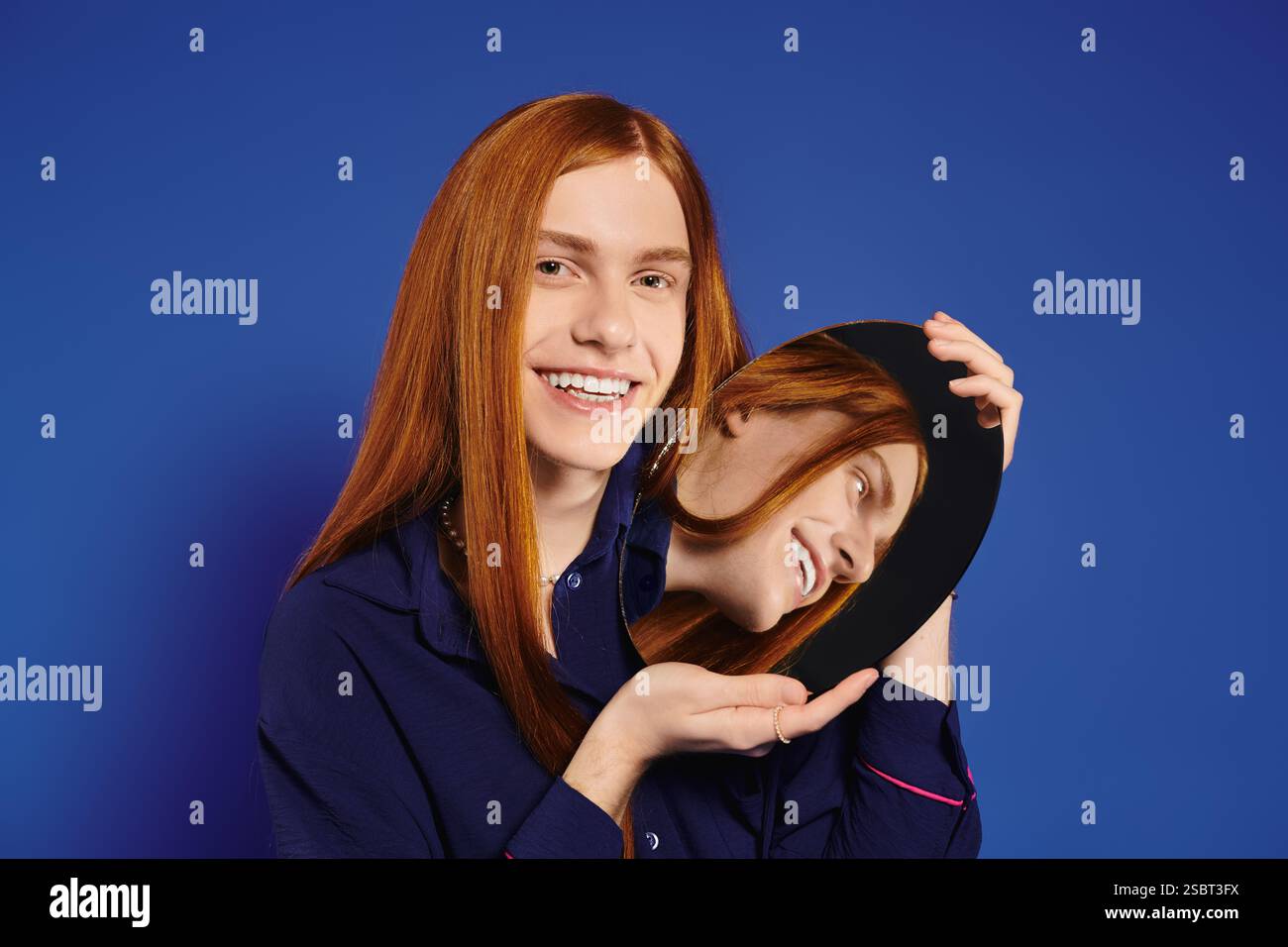 A young queer man with long red hair smiles, holding a mirror that ...