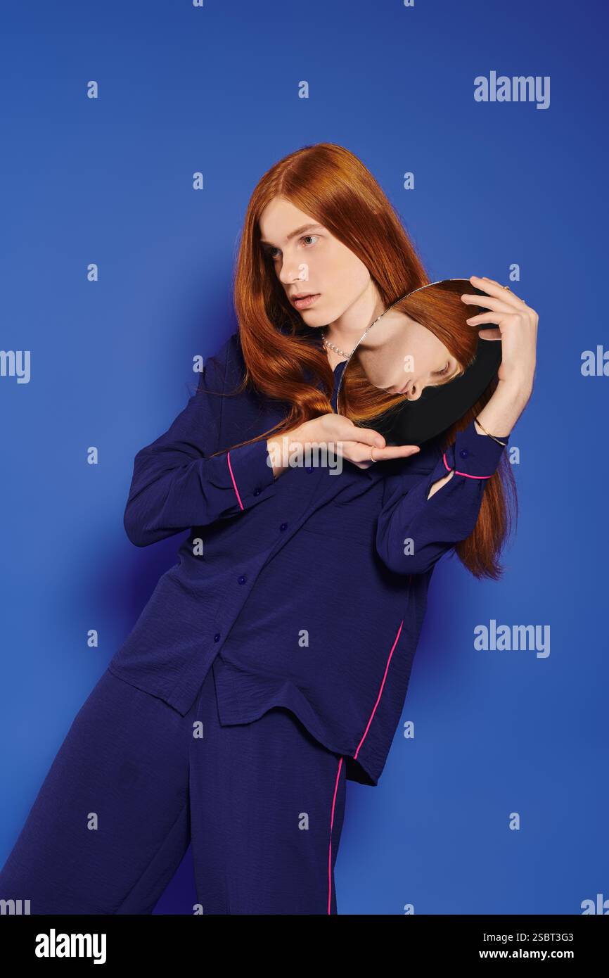 Stylish young queer man with long red hair holds a circular mirror ...