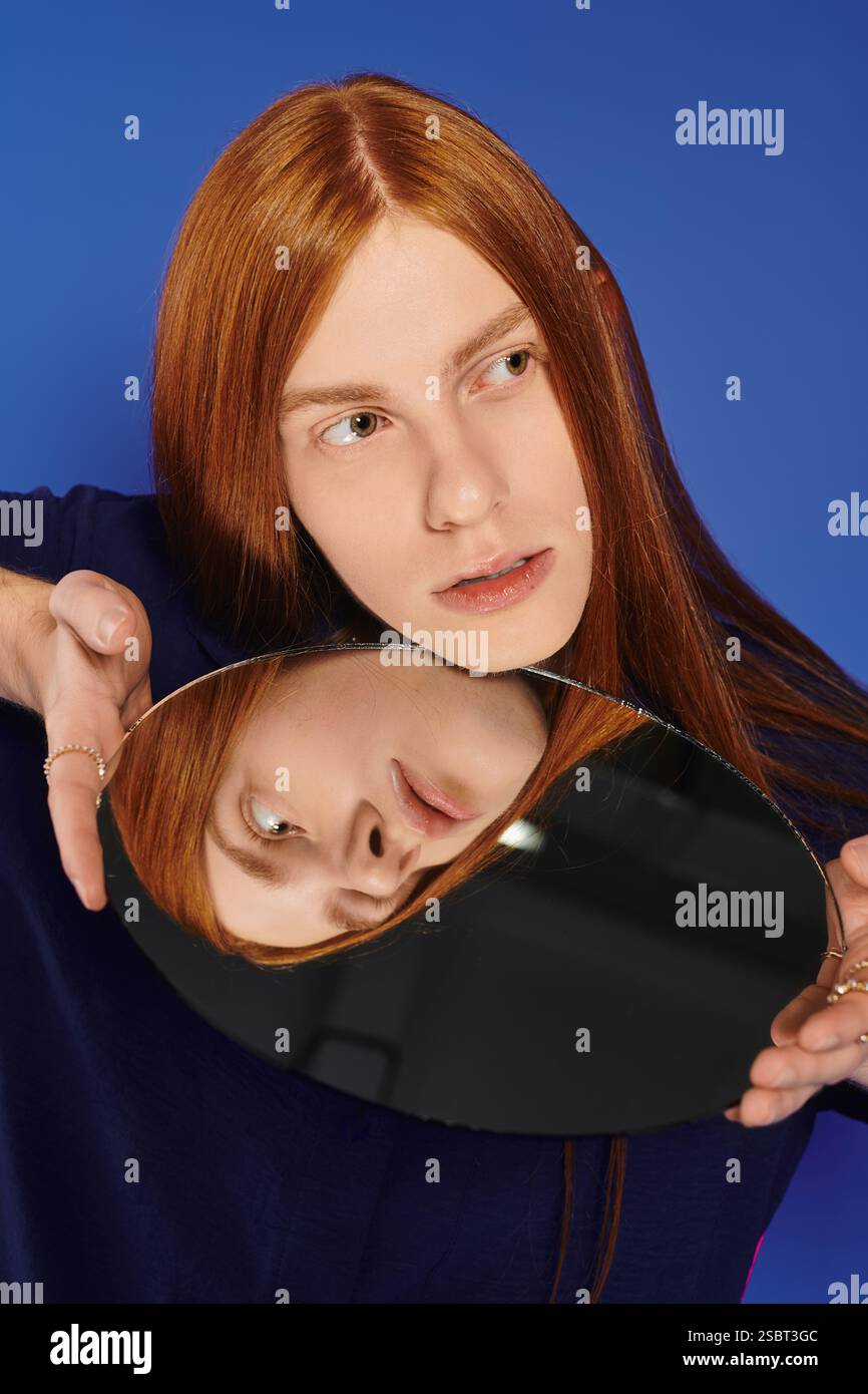 A young queer man with long red hair gazes thoughtfully into a mirror ...