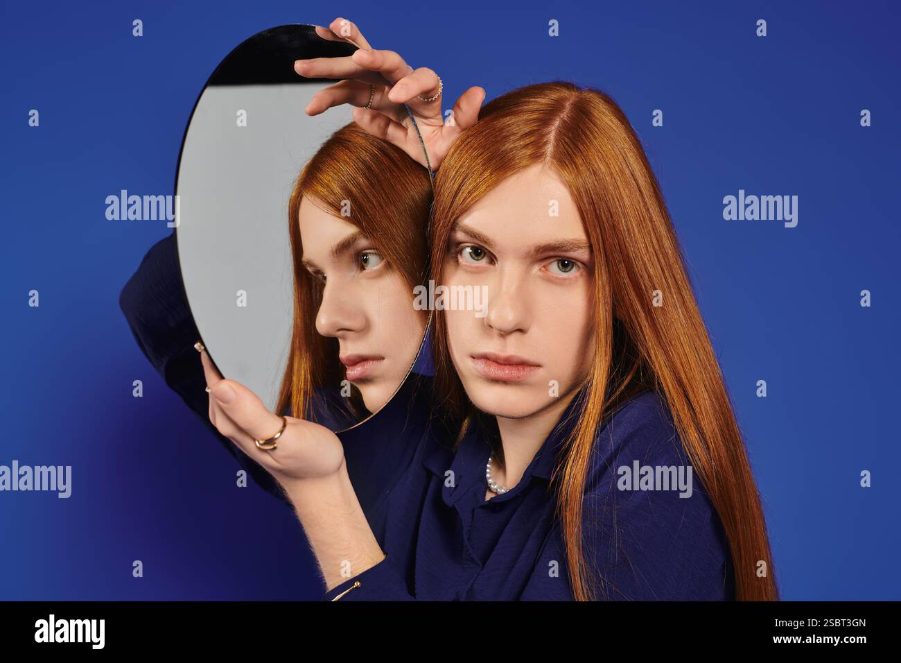 A young queer man with long red hair stares at his reflection in a blue ...