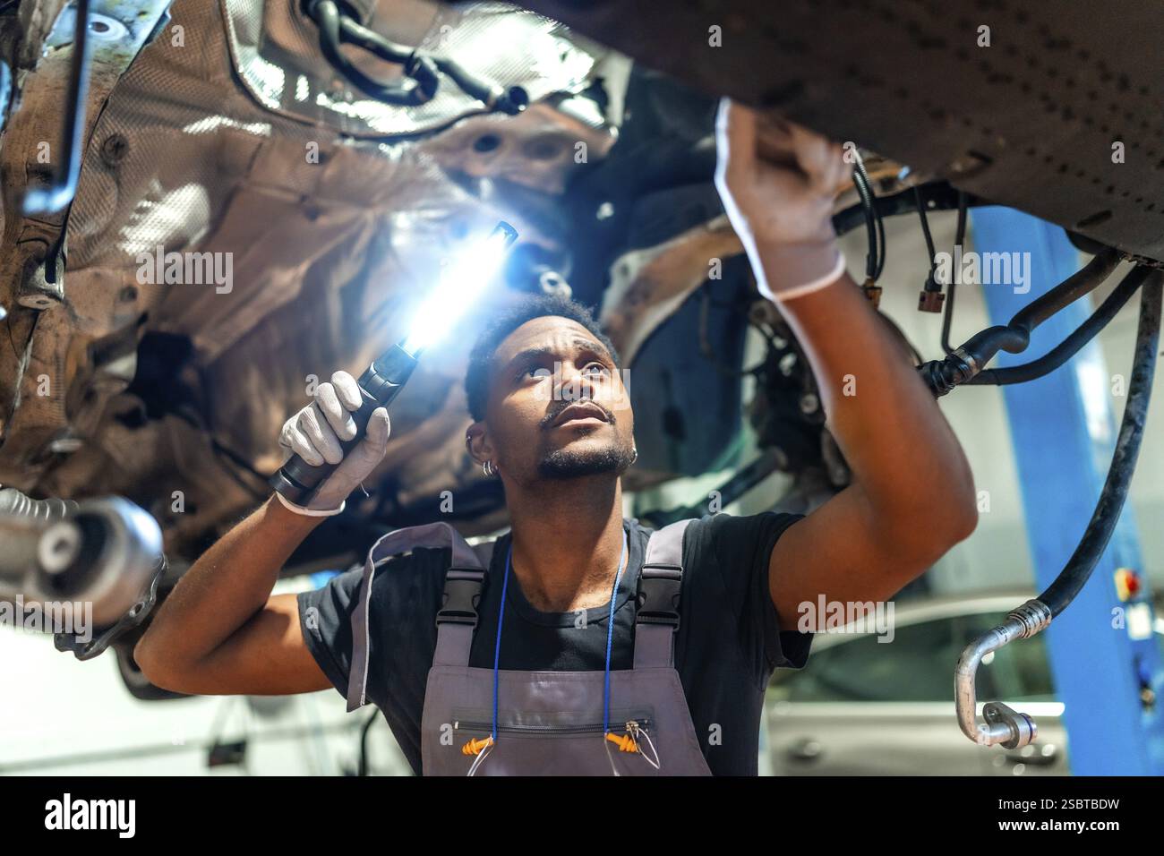 Young mechanic using flashlight, inspecting underside of vehicle raised ...