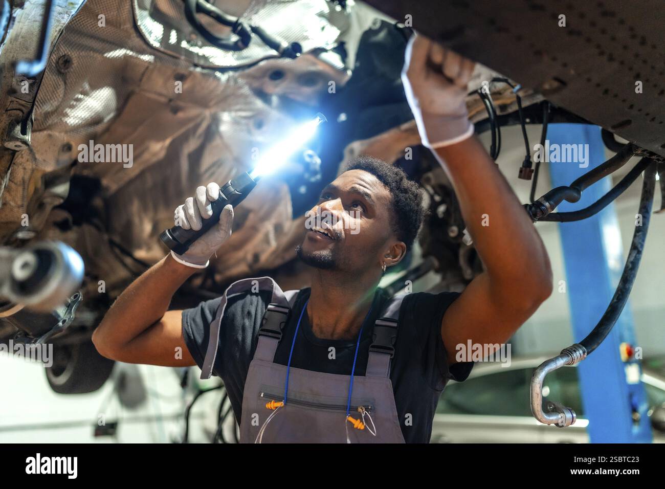 Young mechanic using flashlight, inspecting underside of vehicle raised ...