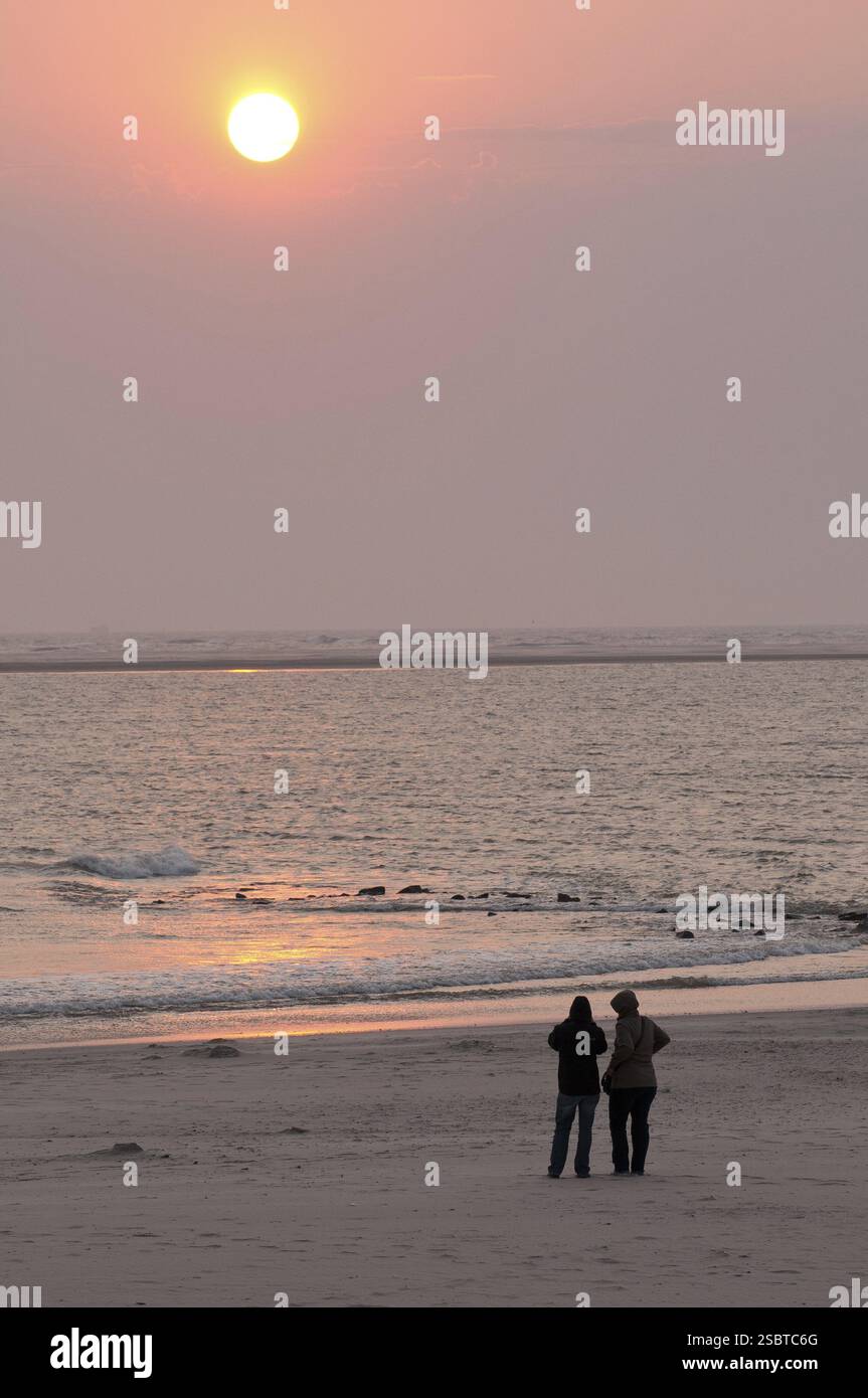 Two people enjoy the sunset at the sea, warm colours fill the horizon, Borkum, north sea, east ...