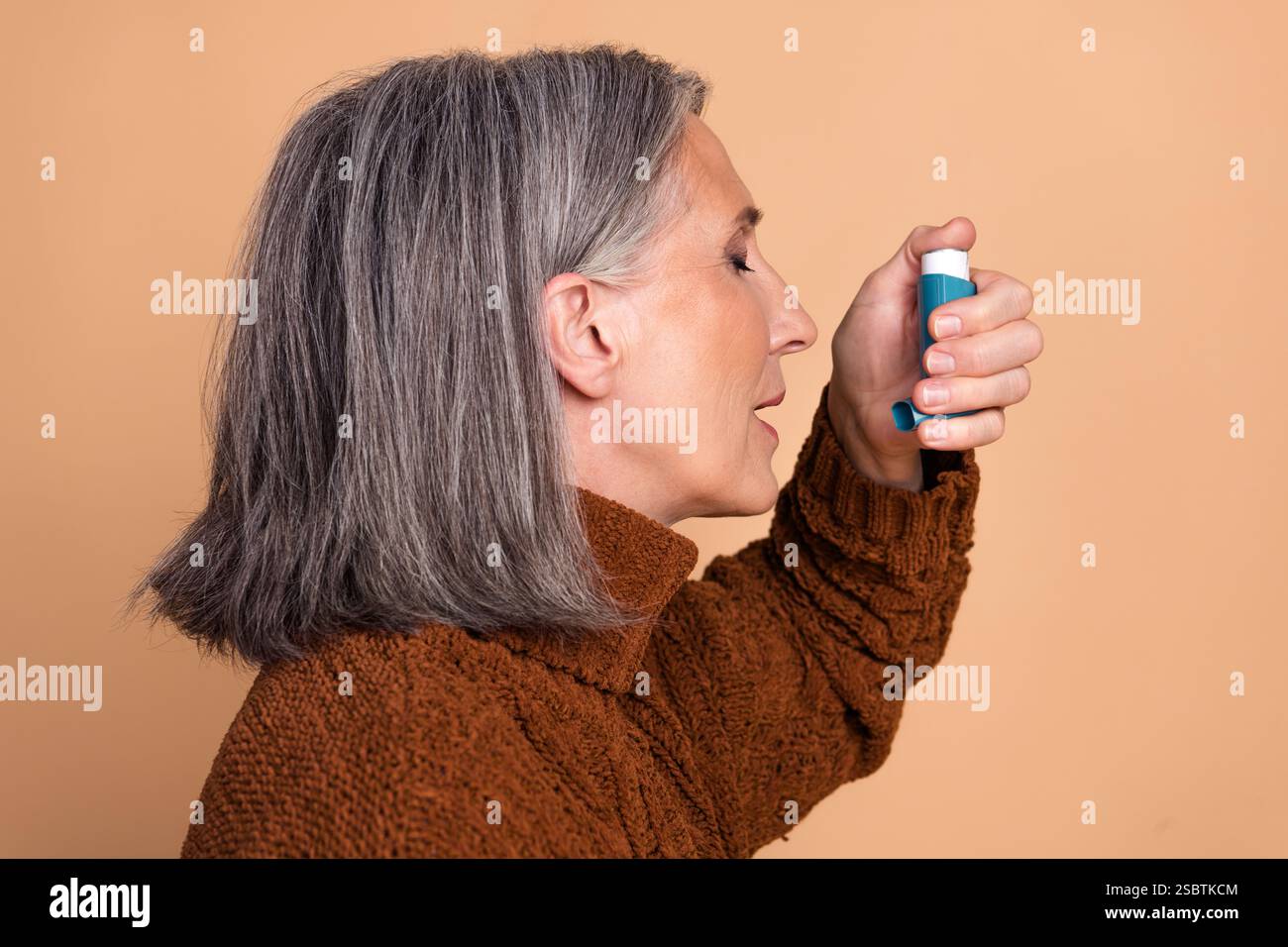 Elderly woman using an inhaler against a beige background wearing a ...