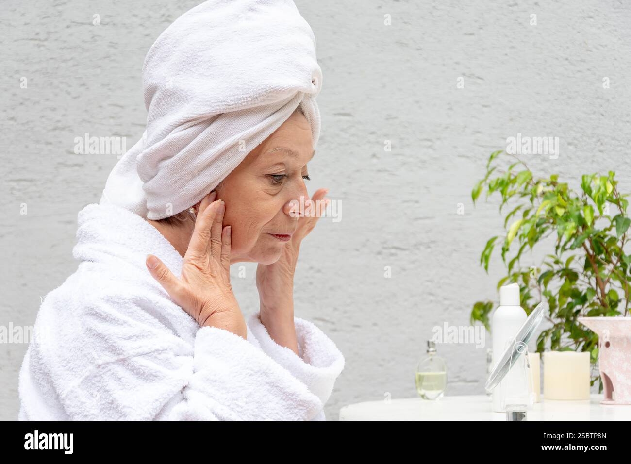 An older woman in a robe takes care of her skin in a home environment ...