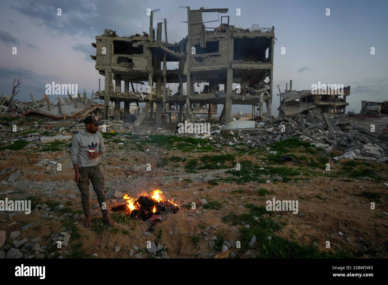 A man stands by a fire next to a destroyed house in an area littered ...