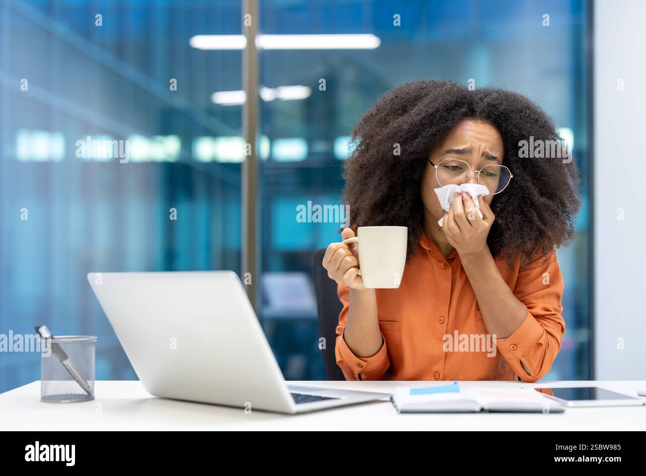 Sick and cold woman at workplace inside office. Employee holding cup of ...