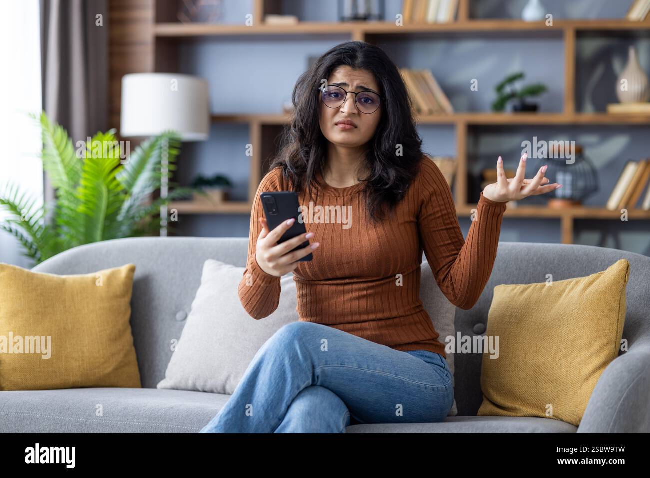 Portrait of upset and disappointed young Muslim woman sitting on sofa at home, holding phone and ...