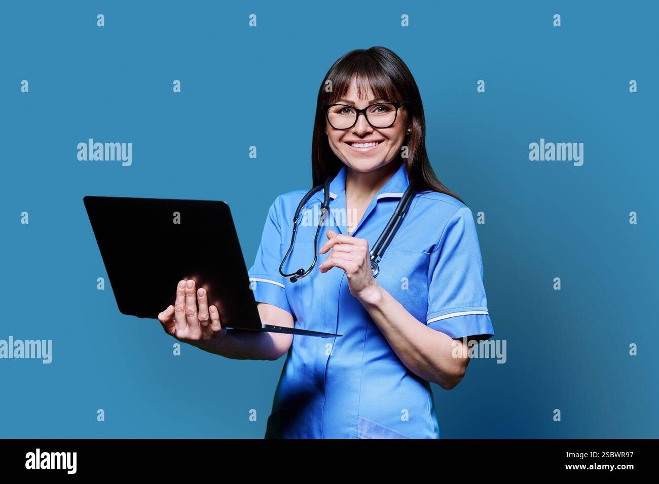 Female nurse holding laptop, looking at camera on blue studio ...