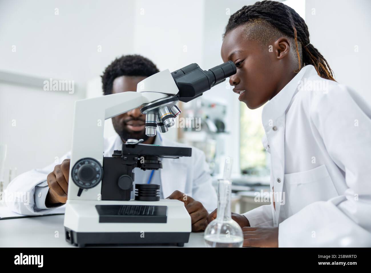 Dark-skinned schoolgirl in a lab coat looking into microscope and ...