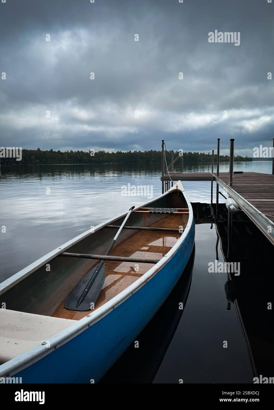 A blue canoe tied to a dock floats on a calm grey lake, dark ominous ...