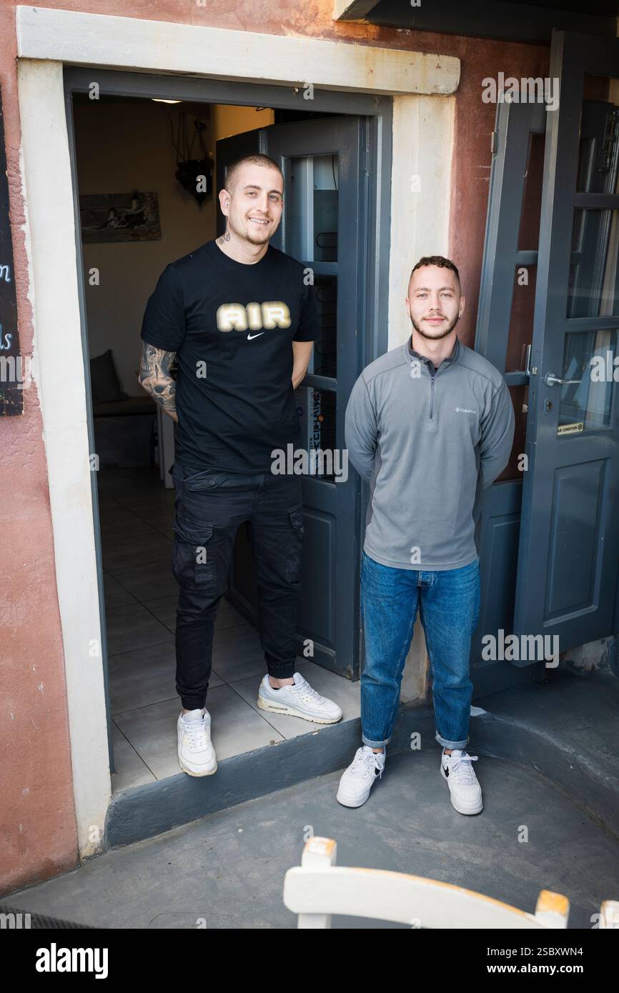 Two young Greek men stand in the entrance of their café in the village ...