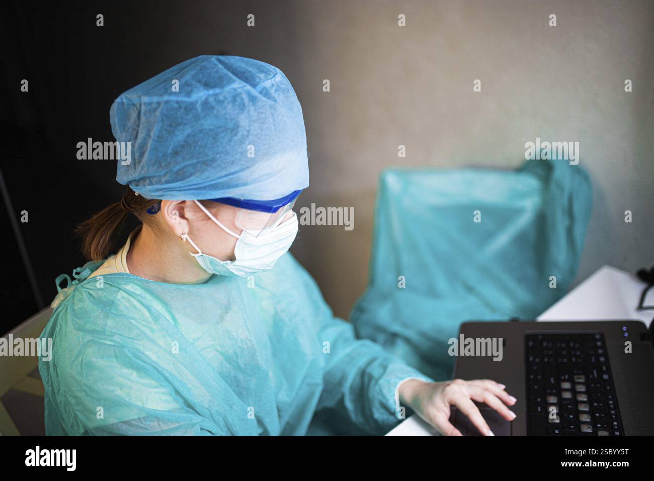 A medical professional in blue gear typing on a laptop at a workstation ...