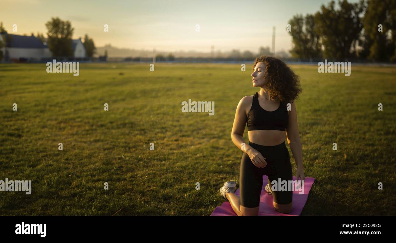 A woman kneels on an exercise mat in a grassy field as the sun sets ...