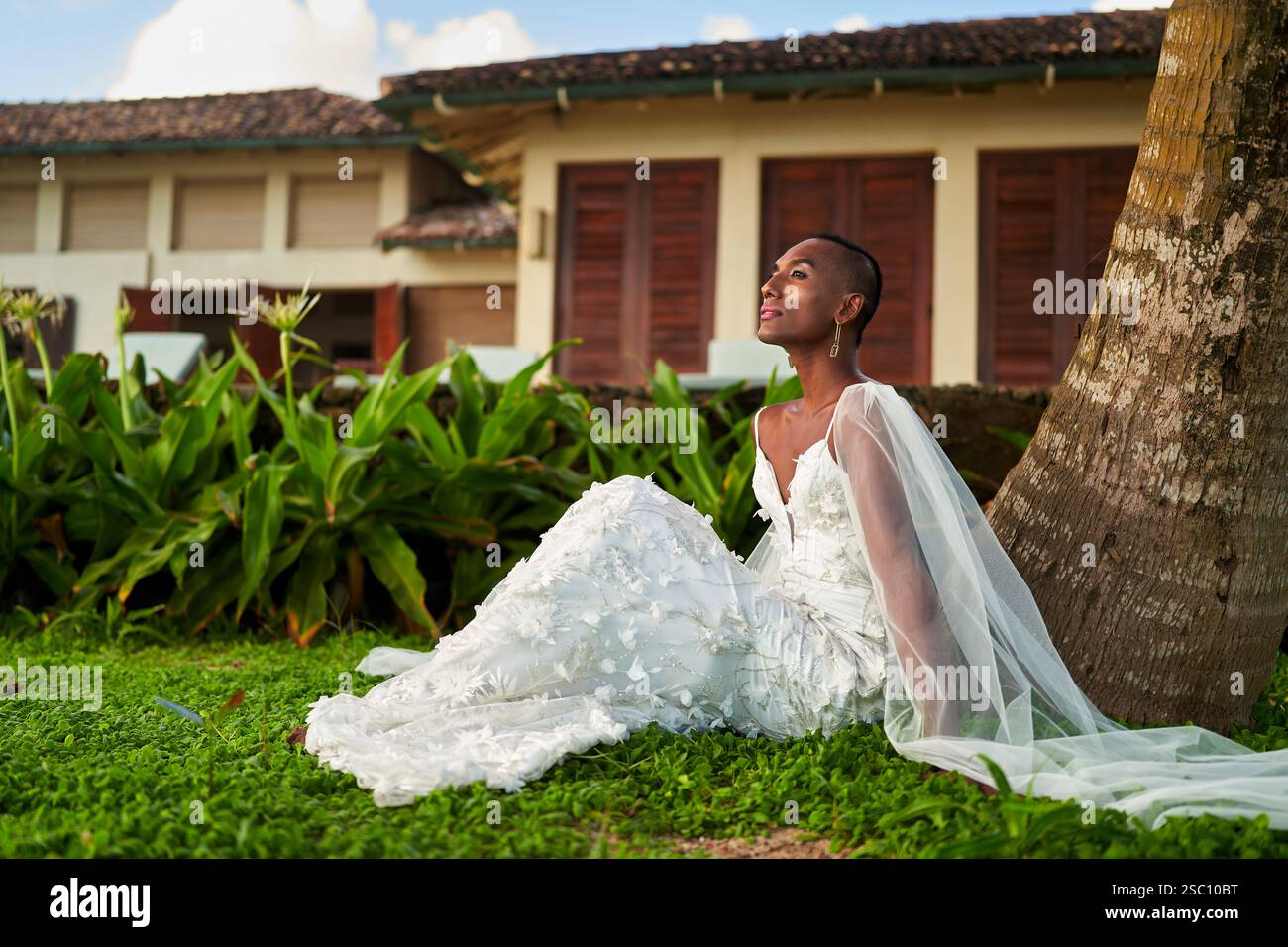 Gender fluid person in white wedding dress sits by palm tree, embracing ...