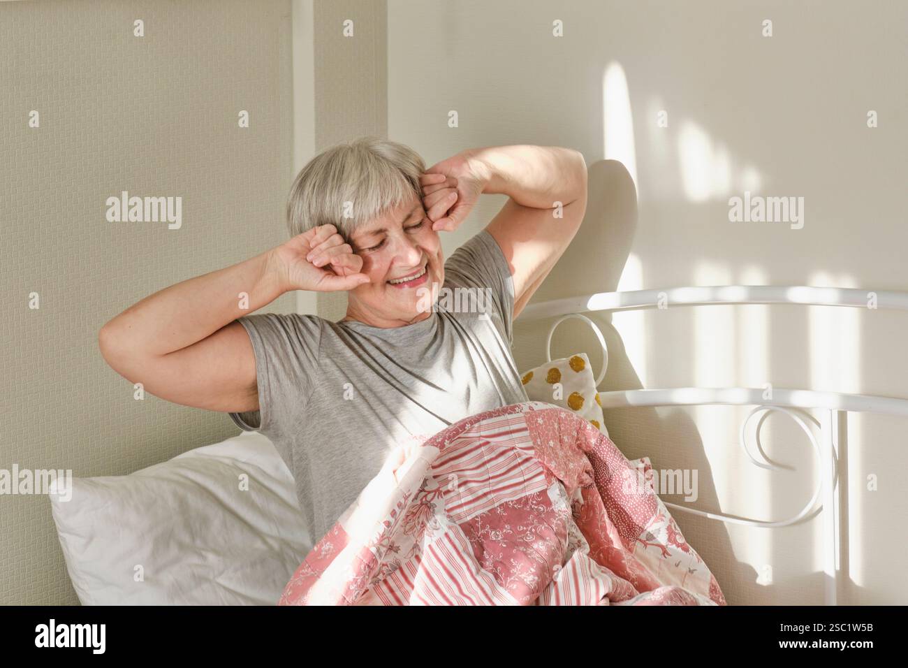 Happy senior woman stretching and rubbing her eyes while waking up in cozy bedroom. Sunlight ...