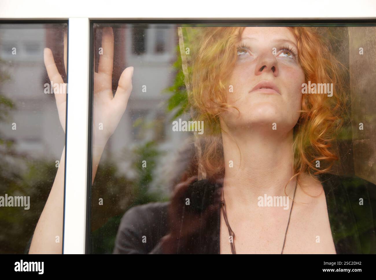 A melancholic redhead woman gazes longingly through a glass window, her ...