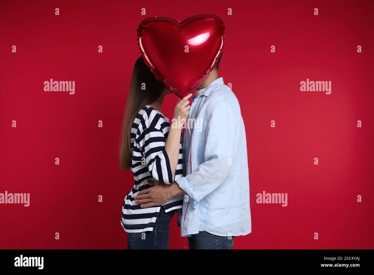 Lovely couple kissing behind heart shaped balloon on red background ...