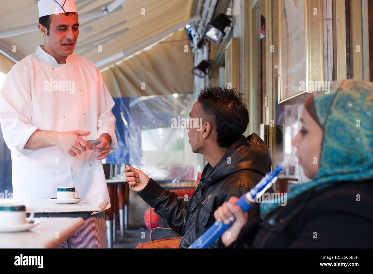 A chef interacts with patrons at a cafe in Istanbul, Turkey as one enjoys a hookah pipe Stock ...