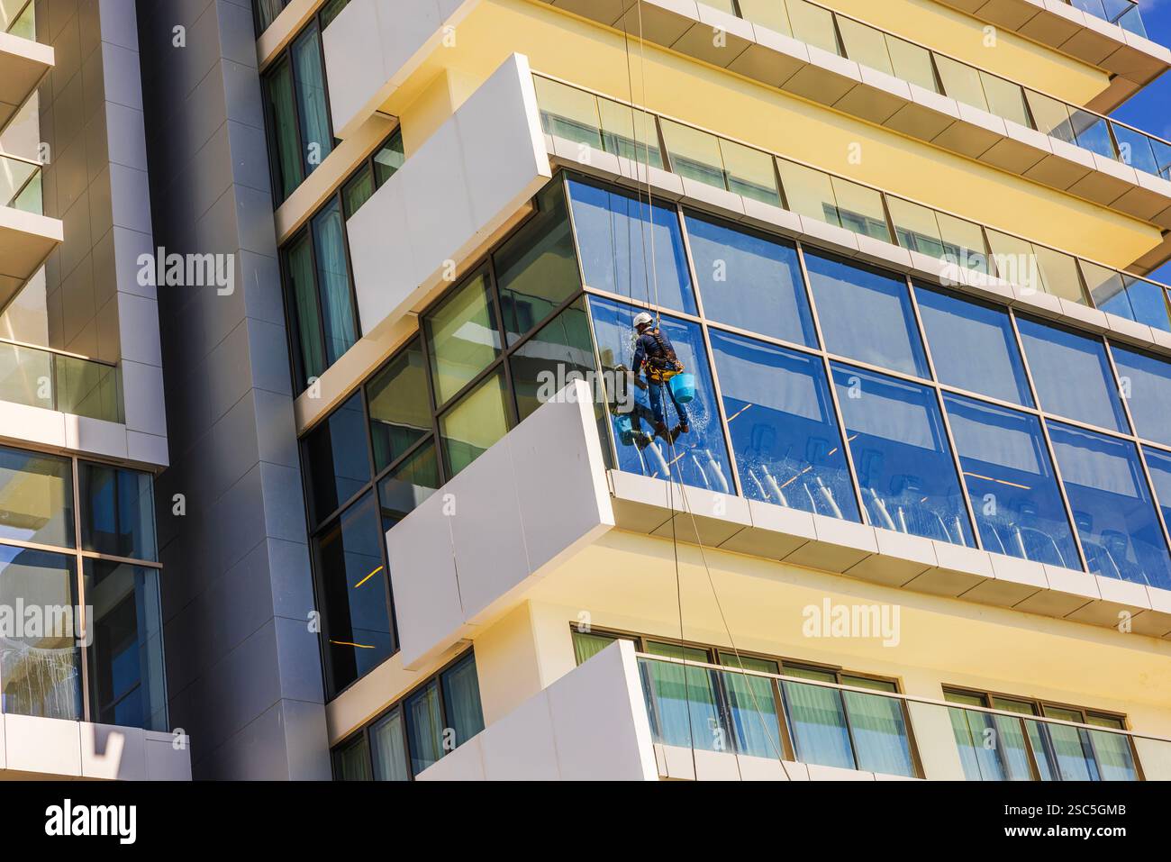 Worker in safety gear cleaning high-rise glass windows using ropes on ...
