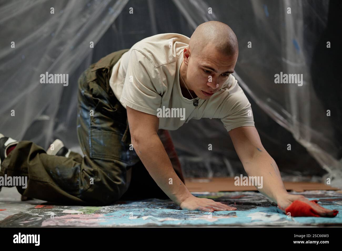 Modern male artist with buzz cut kneeling down on floor to clean working surface stained with ...