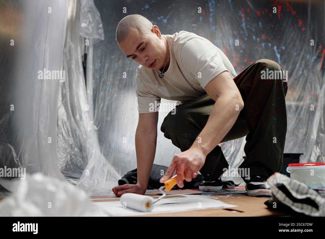 Low angle shot of male artist with buzz cut painting print with roller using paper stencil on ...