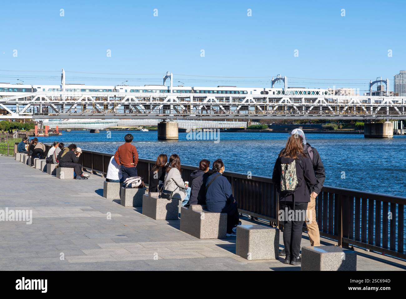 People sitting on the Sumida river walk in Asakusa, looking at a train ...