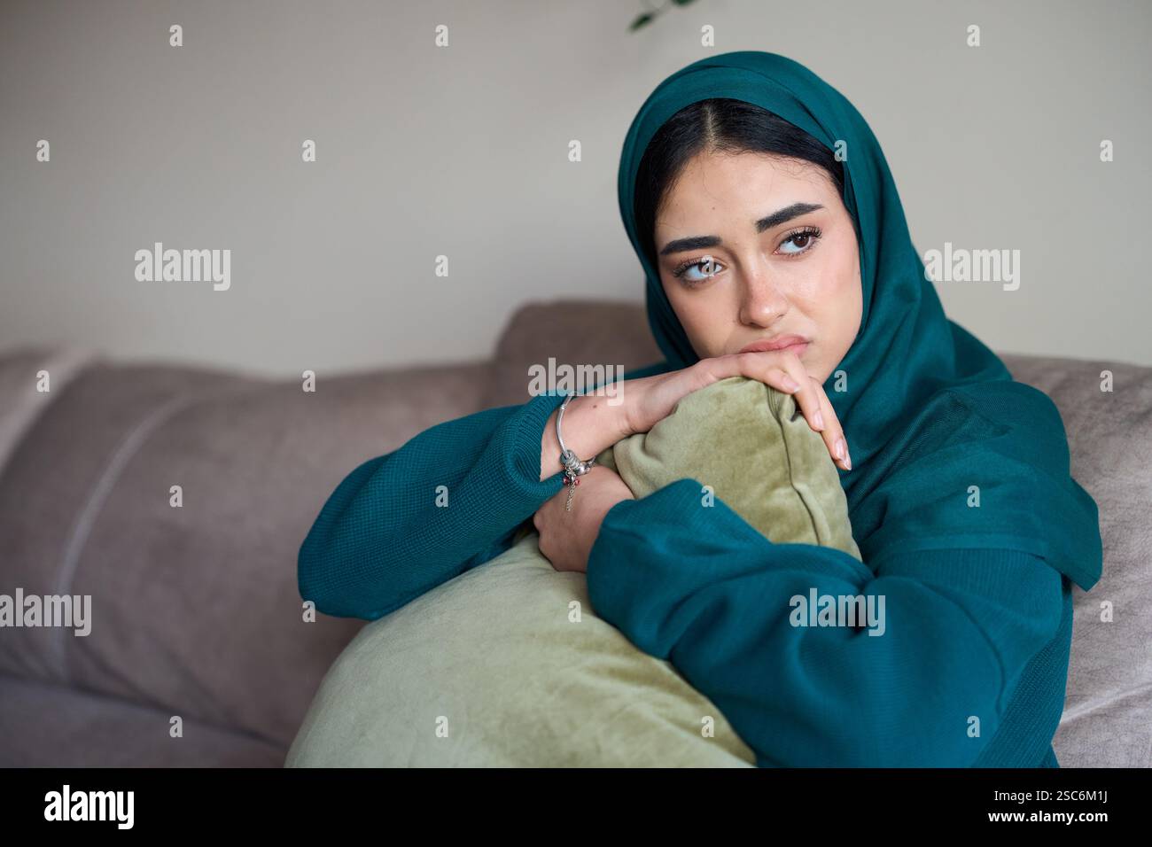 Sad muslim woman wearing hijab sitting on sofa at home hugging pillow ...
