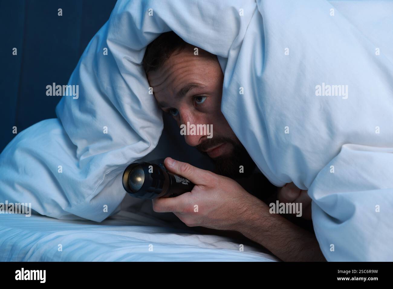 Fear of darkness. Scared man with blanket and flashlight on bed in room ...