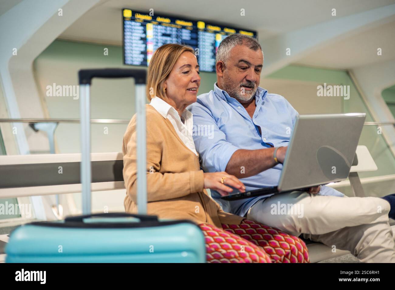 Senior couple sitting on a bench at the airport terminal using a laptop while waiting for their ...