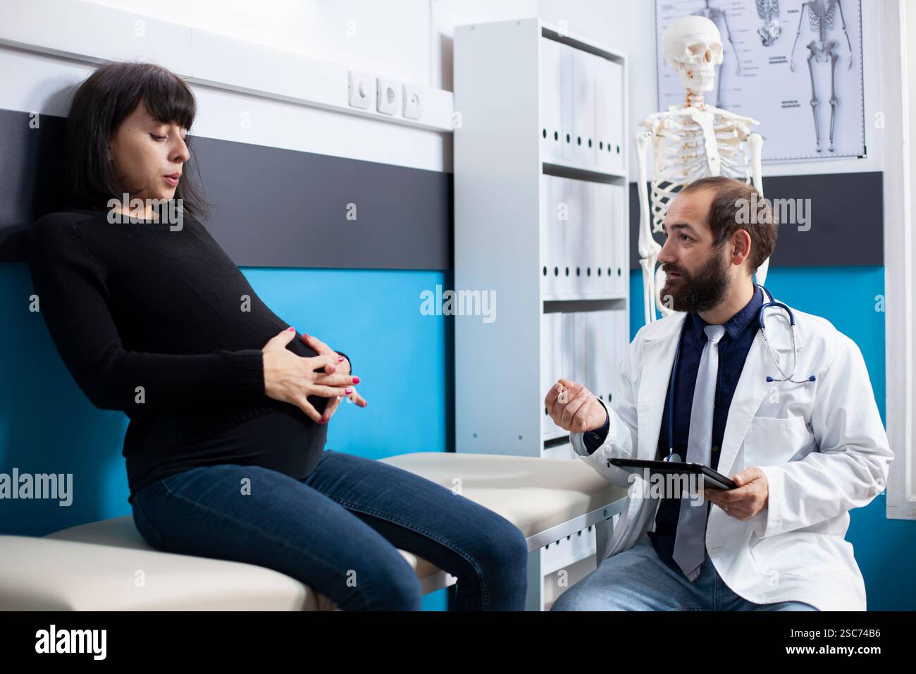 Pregnant patient gently stroking belly bump as young doctor with tablet ...