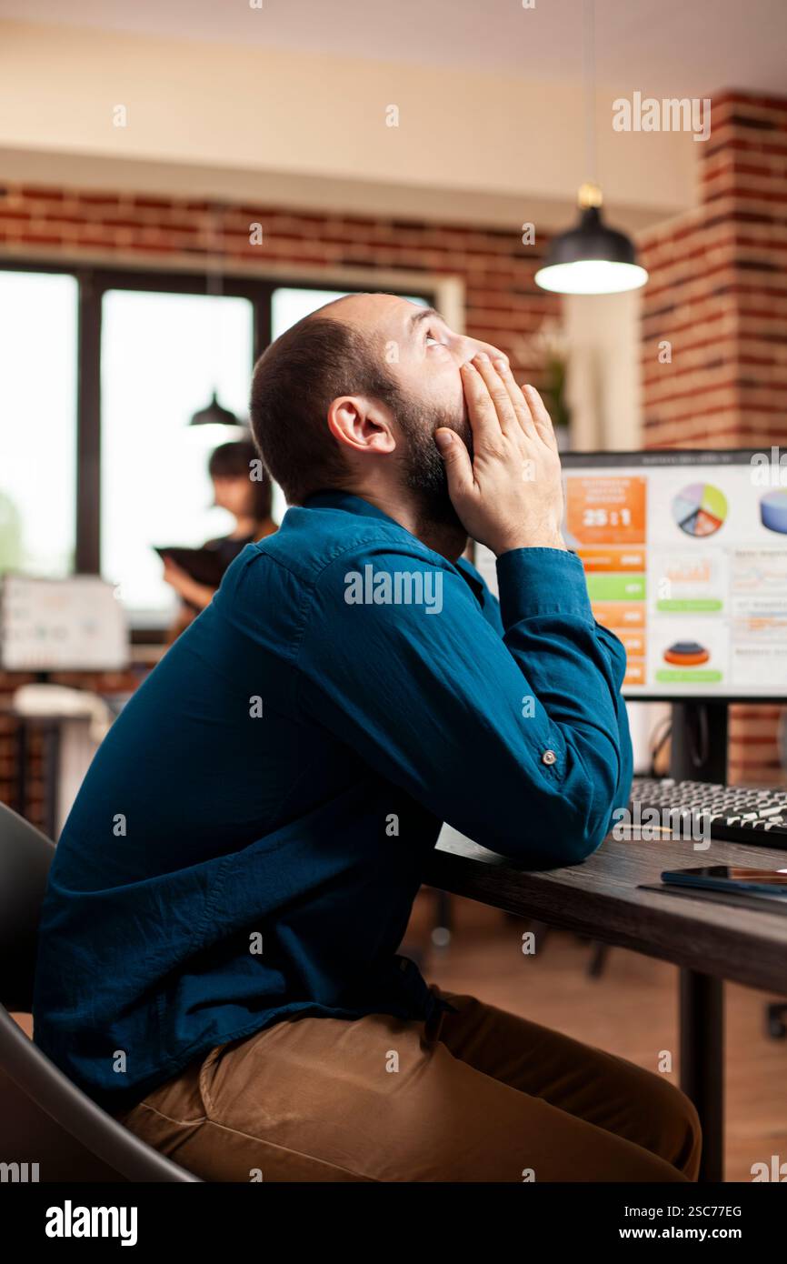 Young male employee seated at office desk, rubbing his face from ...