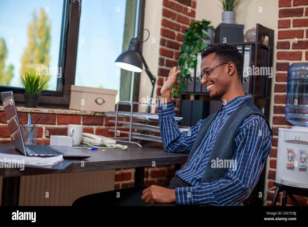 Young male employee wearing wireless earphones waves at his laptop ...