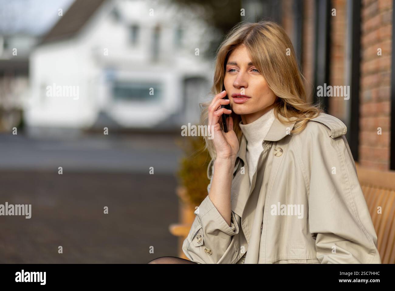 A young woman wearing a light trench coat sits on a bench, engaged in a ...