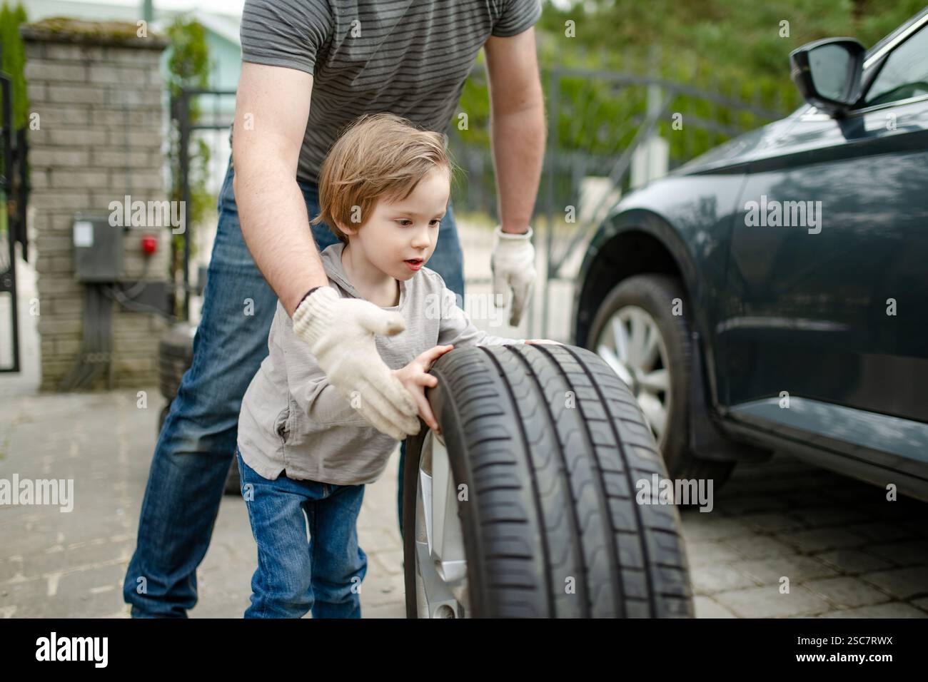 Cute little boy helping his father to change car wheels at their backyard. Father teaching his ...