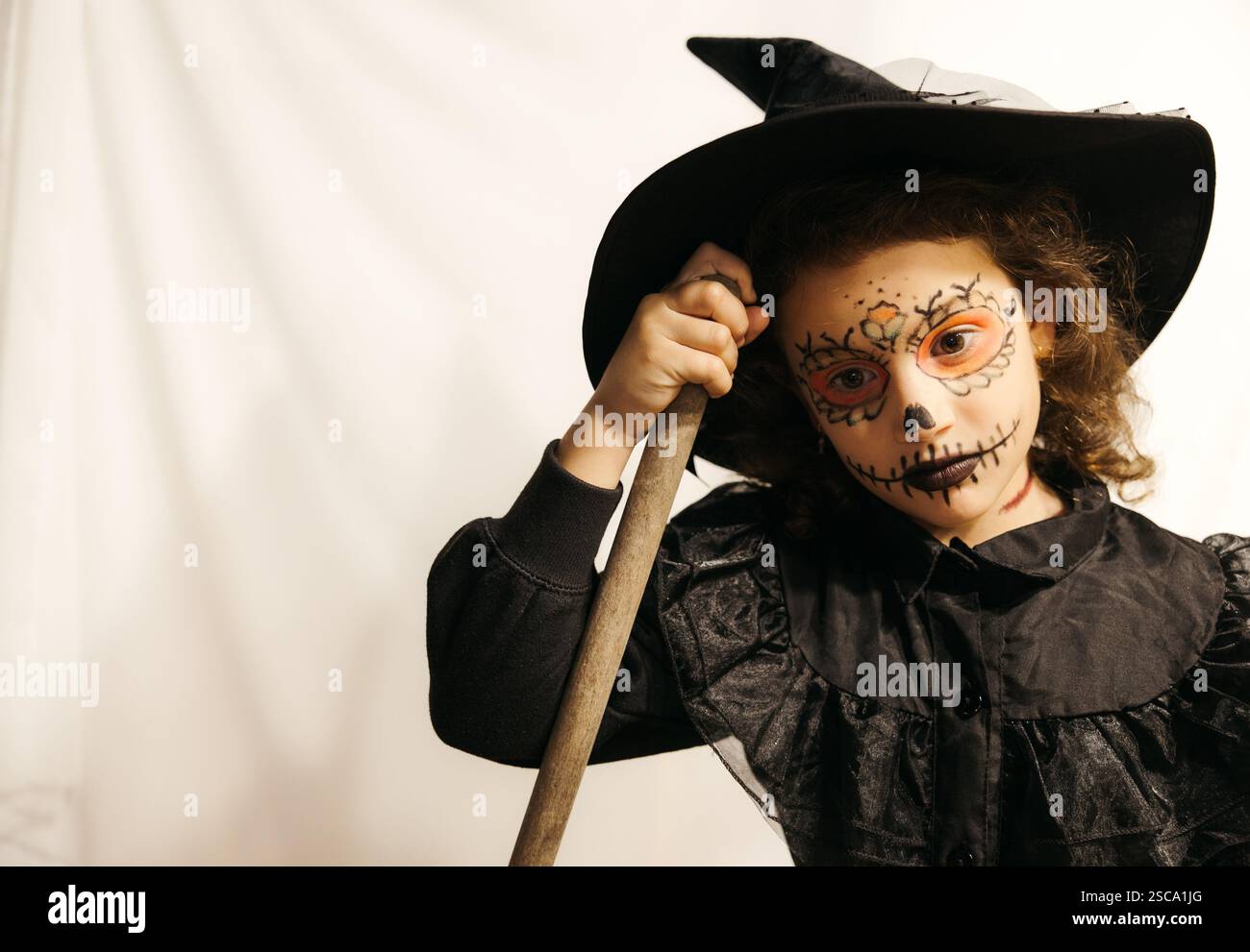 A young girl dressed as a witch with skull face paint, holding a broom ...