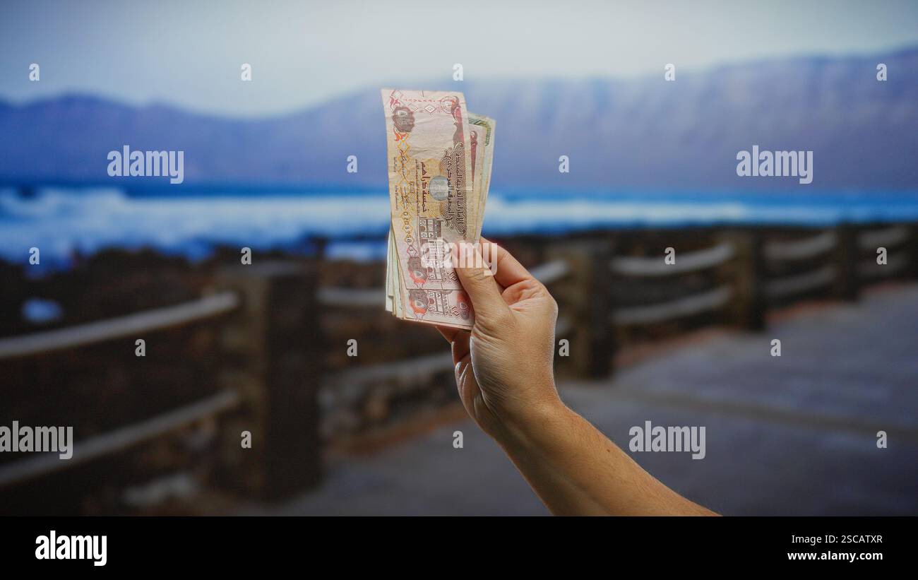 Hand holding uae dirhams against a scenic seaside backdrop featuring a ...