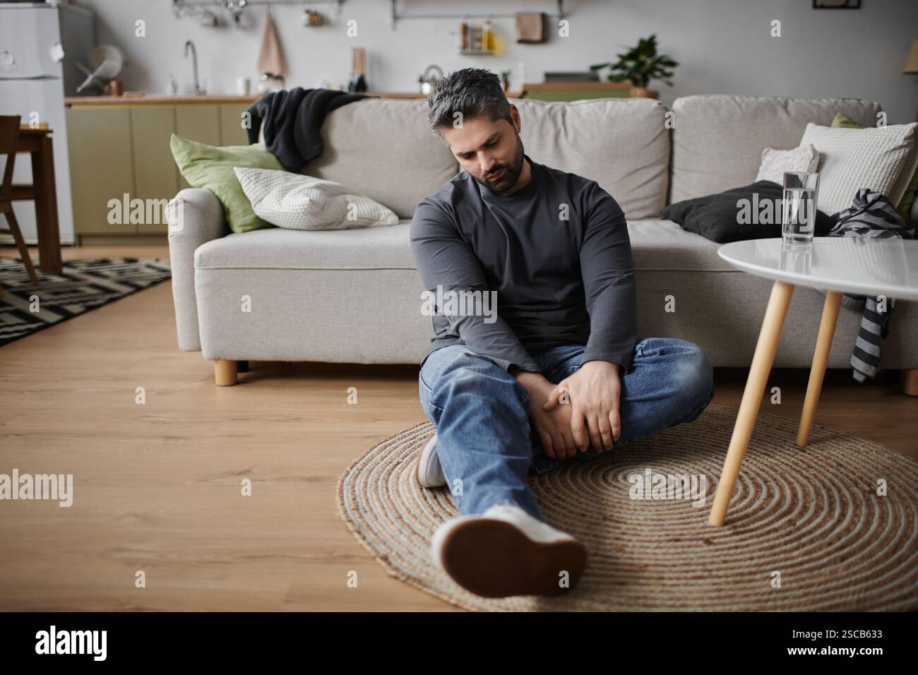 A handsome man sits thoughtfully on the floor of his inviting living space, lost in reflection ...