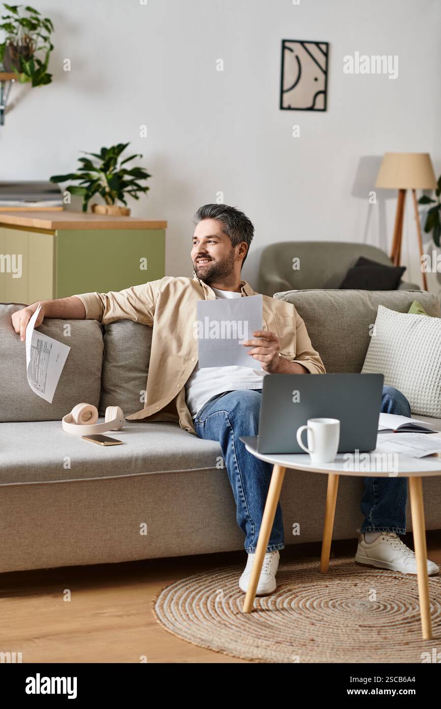 A handsome man sits comfortably on a cozy couch, holding papers and ...