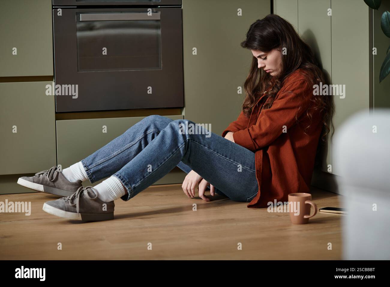 A young woman sits on the kitchen floor, lost in thought, grappling ...
