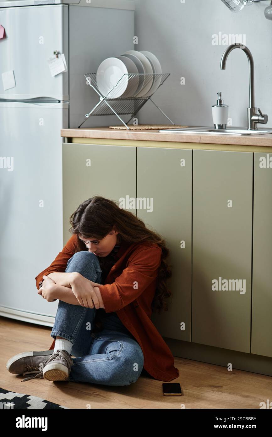 A woman sits alone on the kitchen floor, embracing her knees, lost in ...