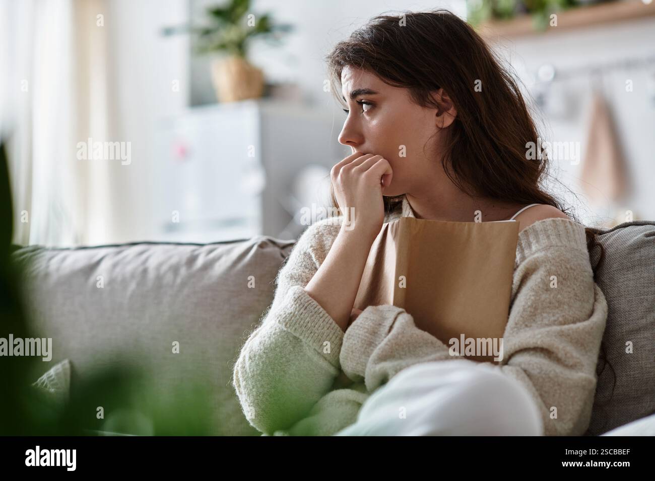A woman sits on a couch, lost in thought, reflecting on her emotions ...