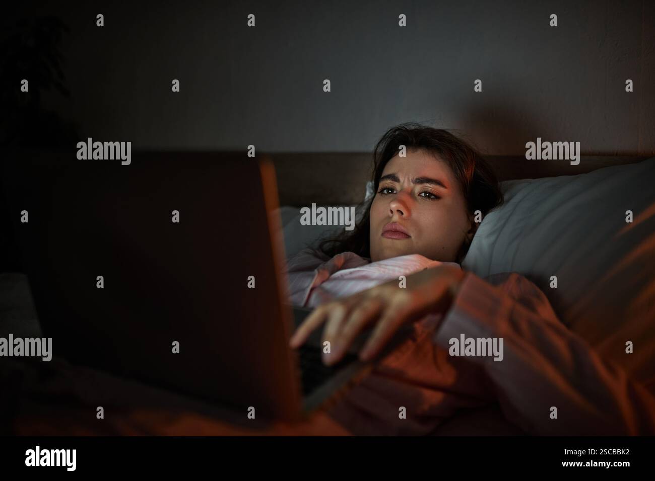 A woman lies in bed, focused on her laptop, reflecting feelings of ...