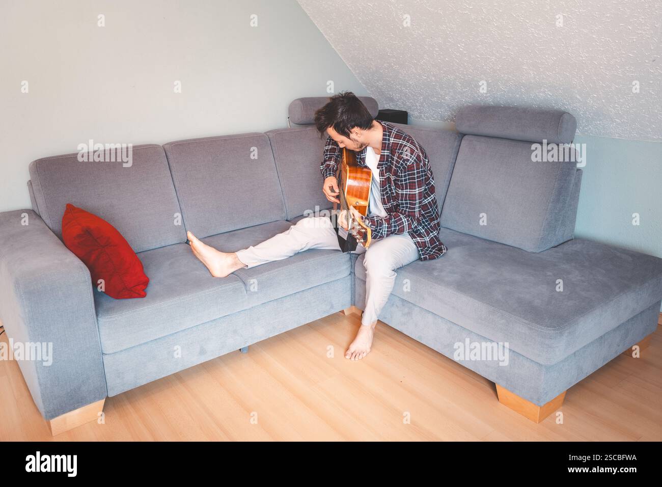 Barefoot musician sits comfortably on a gray couch, strumming an ...
